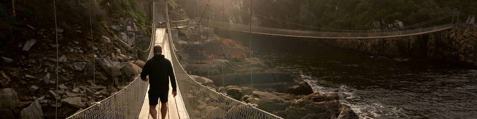 Uma pessoa caminhando sobre uma ponte suspensa sobre um rio, com penhascos rochosos em ambas as margens.