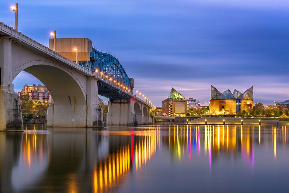 Uma ponte sobre um rio calmo, com reflexos de luzes, um edifício moderno com fachada de vidro e um horizonte urbano ao fundo.