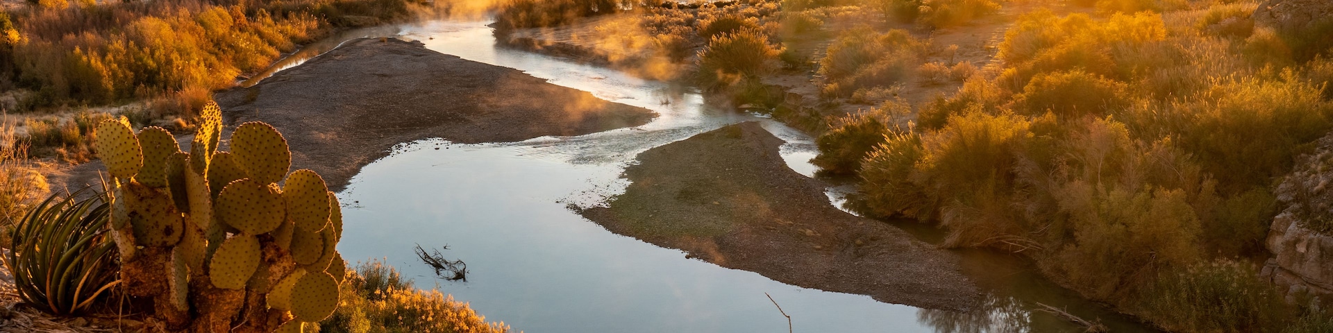 Paisagem desértica com um rio, cactos e montanhas ao fundo.