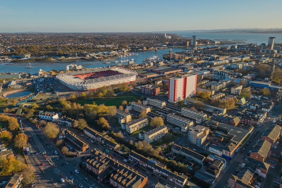 Vista aérea de uma cidade com um estádio grande, um rio e um edifício alto.