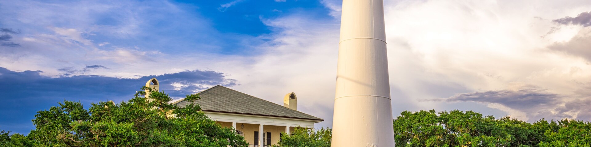 Um farol branco com varanda, uma casa grande com alpendre e árvores verdes exuberantes.
