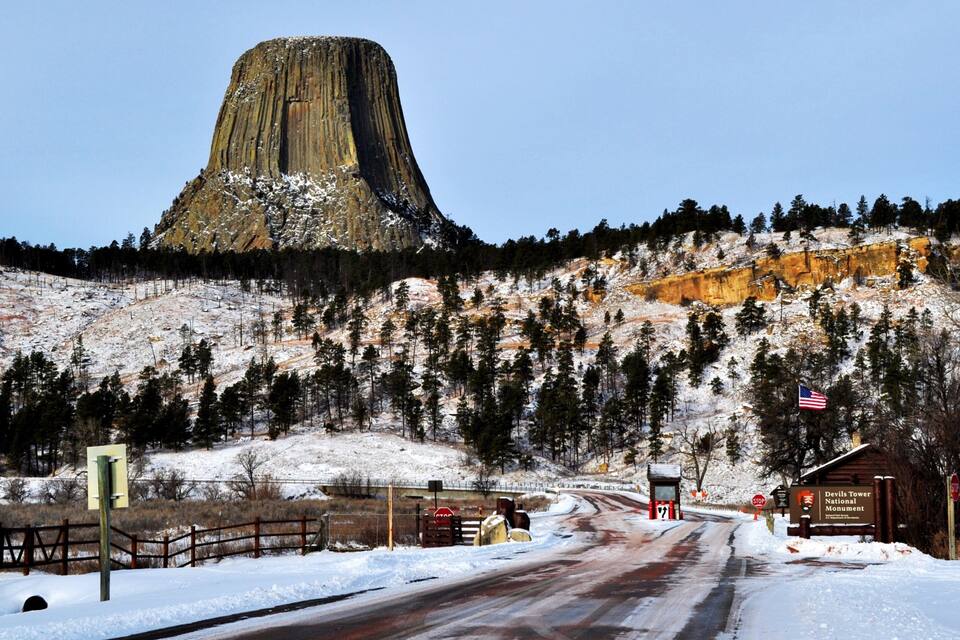 Uma estrada coberta de neve que leva ao Monumento Nacional Devils Tower, com uma formação rochosa imponente ao fundo.
