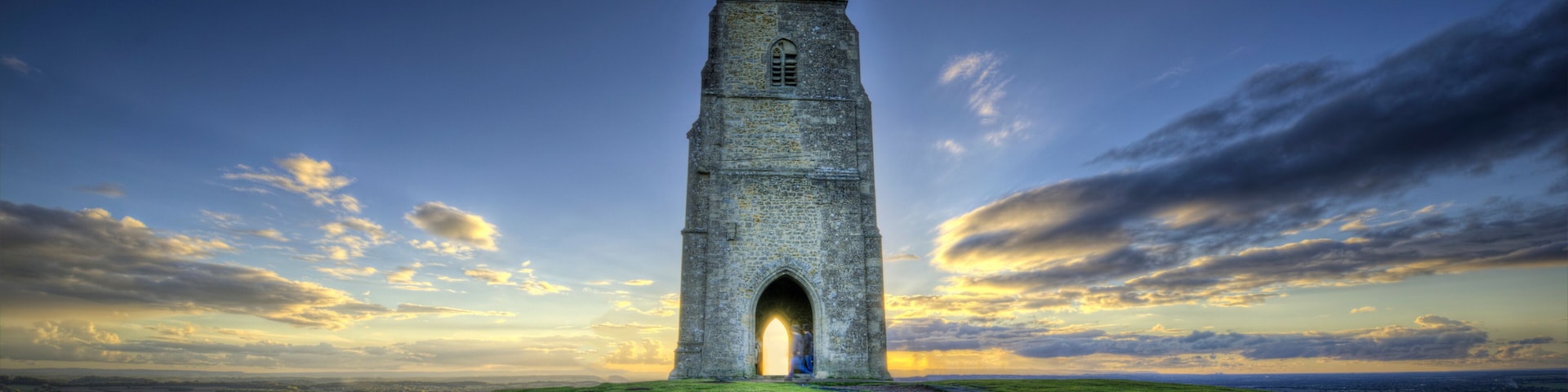 Uma torre com entrada em arco, cercada por um campo gramado e um caminho de cascalho, sob um céu parcialmente nublado.