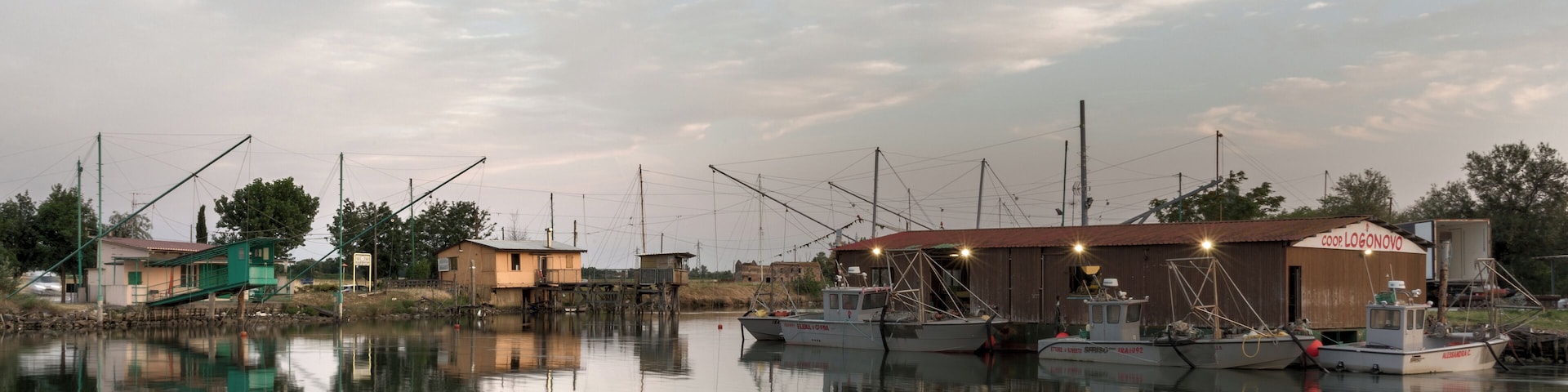 Um porto tranquilo com vários barcos de pesca atracados, um pequeno edifício de madeira e um céu nublado.
