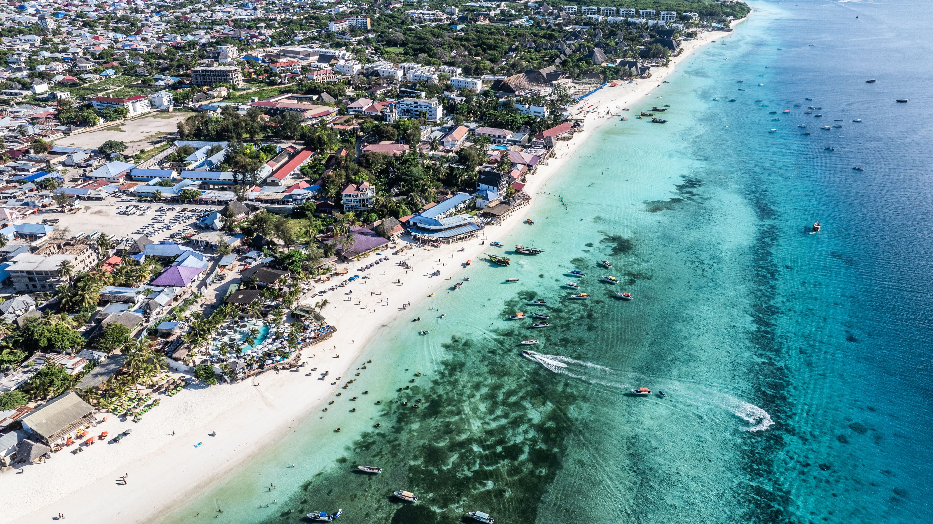 Vista aérea de uma cidade costeira com uma praia, águas turquesa límpidas e diversos barcos.