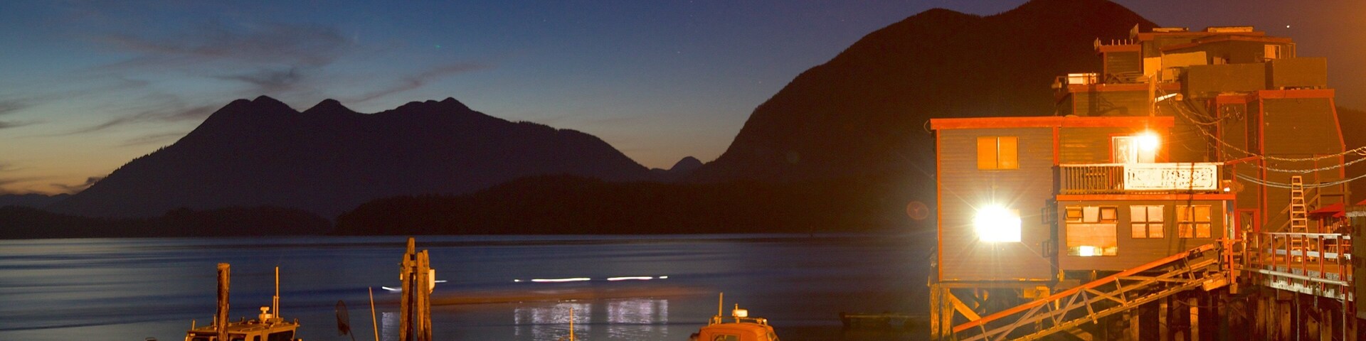Um lago calmo ao entardecer com barcos atracados, um píer de madeira e um edifício bem iluminado com montanhas ao fundo.