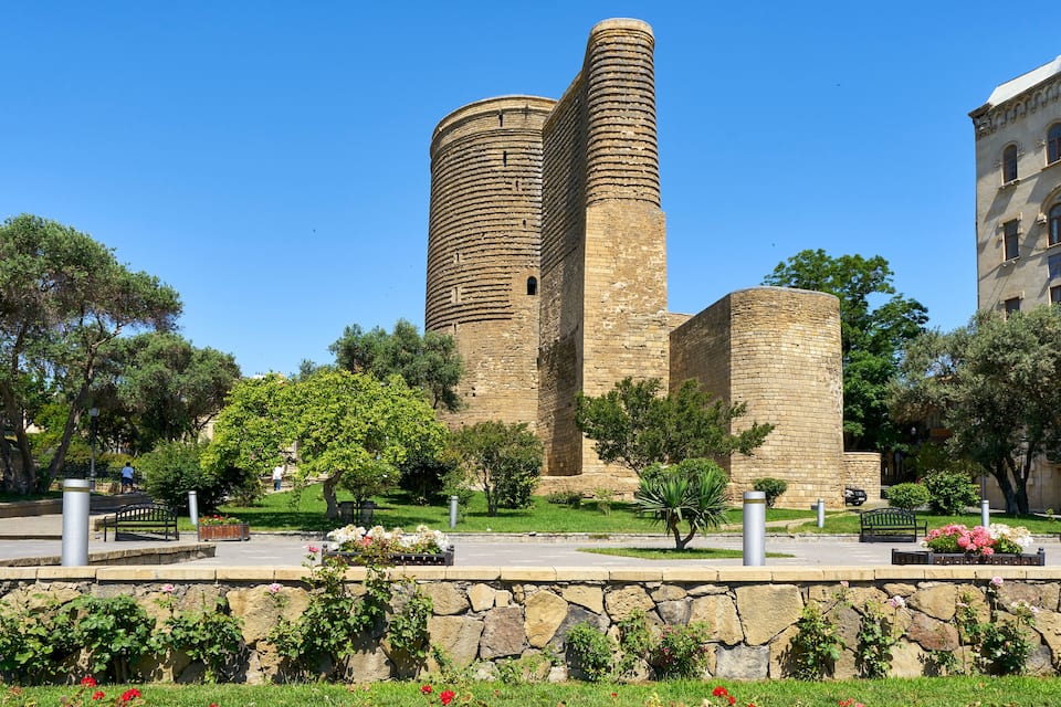 Uma torre de pedra histórica com base cilíndrica e topo quadrado, rodeada por um parque com bancos e plantas floridas.