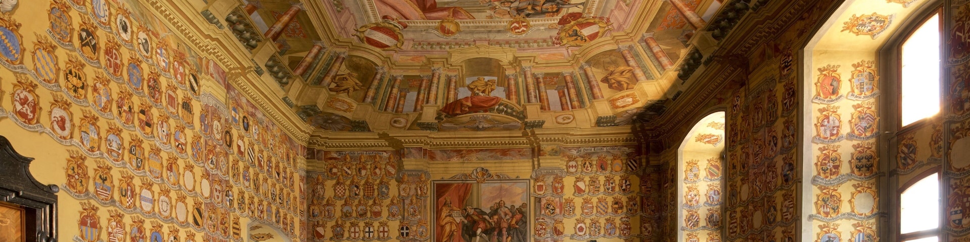 Um salão imponente com tetos ricamente decorados, um grande mural e inúmeros brasões nas paredes.
