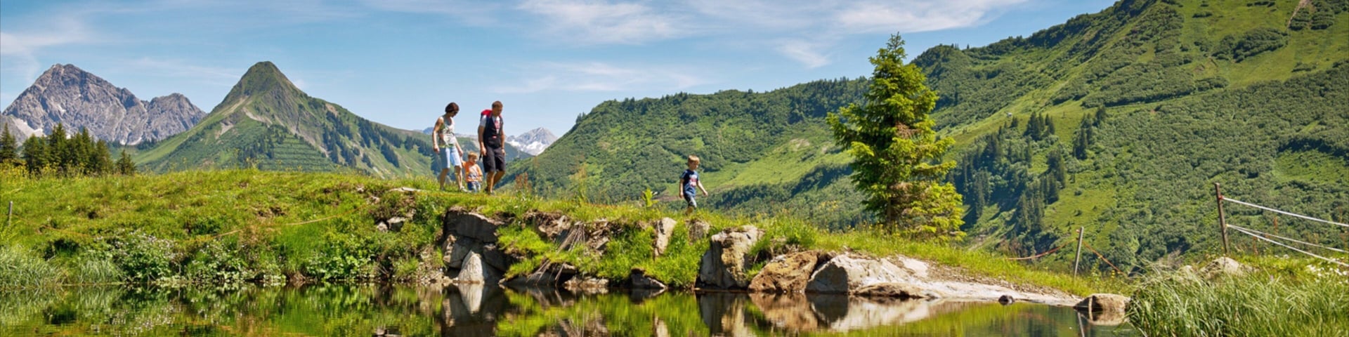 Um grupo de pessoas perto de um lago de montanha com reflexos, rodeado por vegetação exuberante e montanhas.