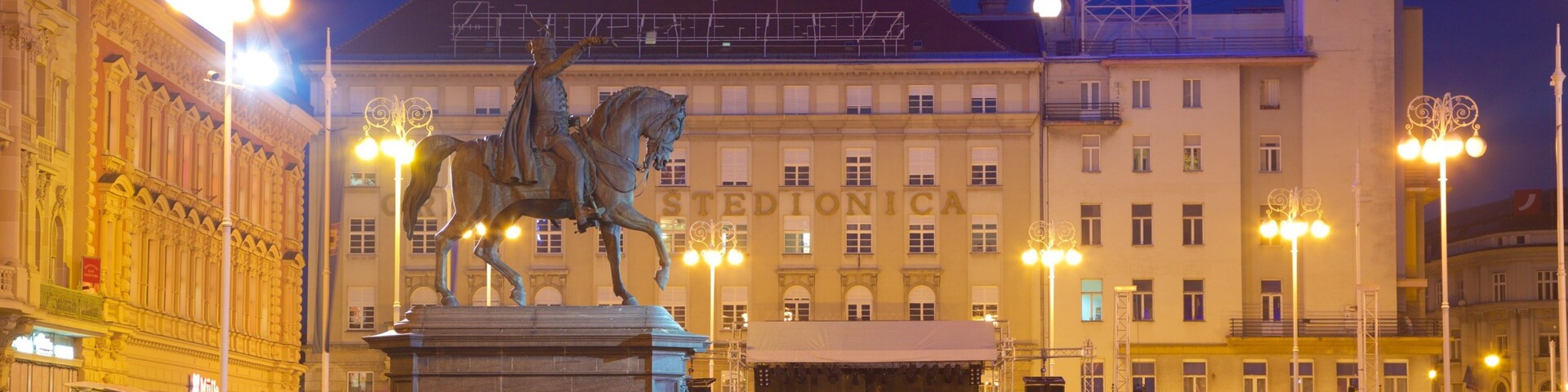Estátua de um homem a cavalo numa praça, com um edifício identificado como 'STEDIONICA' ao fundo.
