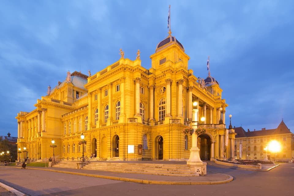 Um edifício imponente, iluminado em amarelo, com arquitetura clássica, incluindo colunas e estátuas, ao entardecer.