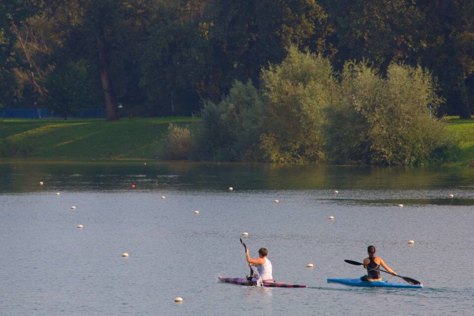 Duas pessoas em caiaques num lago calmo, com boias, rodeadas por árvores e áreas verdes.