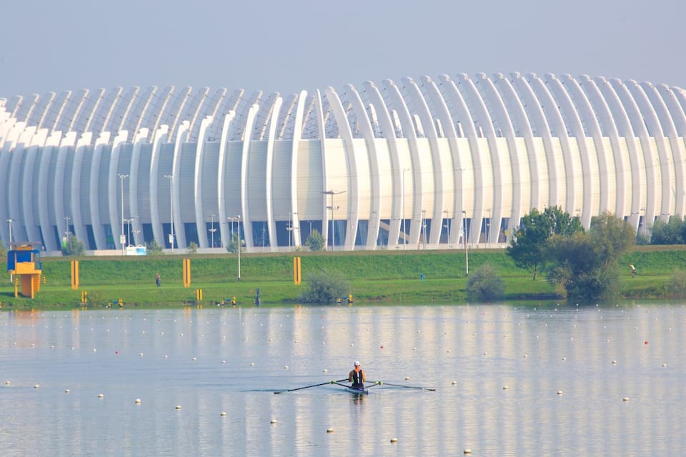 Um estádio moderno com um design de telhado curvo único, um corpo de água ao primeiro plano e algumas pessoas remando.