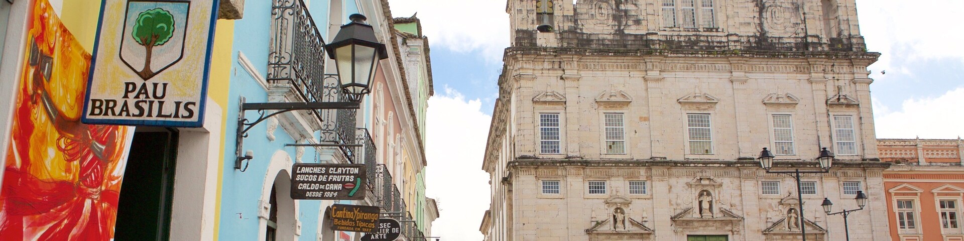 Uma rua colorida com uma placa indicando Pau Brasilis, um edifício histórico com uma cruz no topo, e carros estacionados.