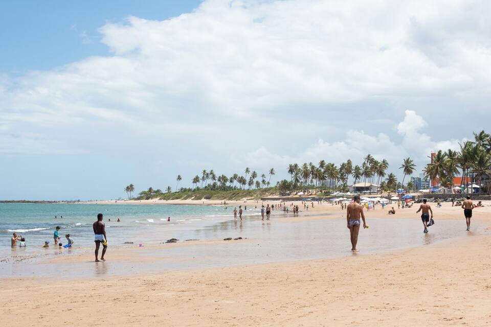 Uma praia com pessoas caminhando, palmeiras e um céu claro.