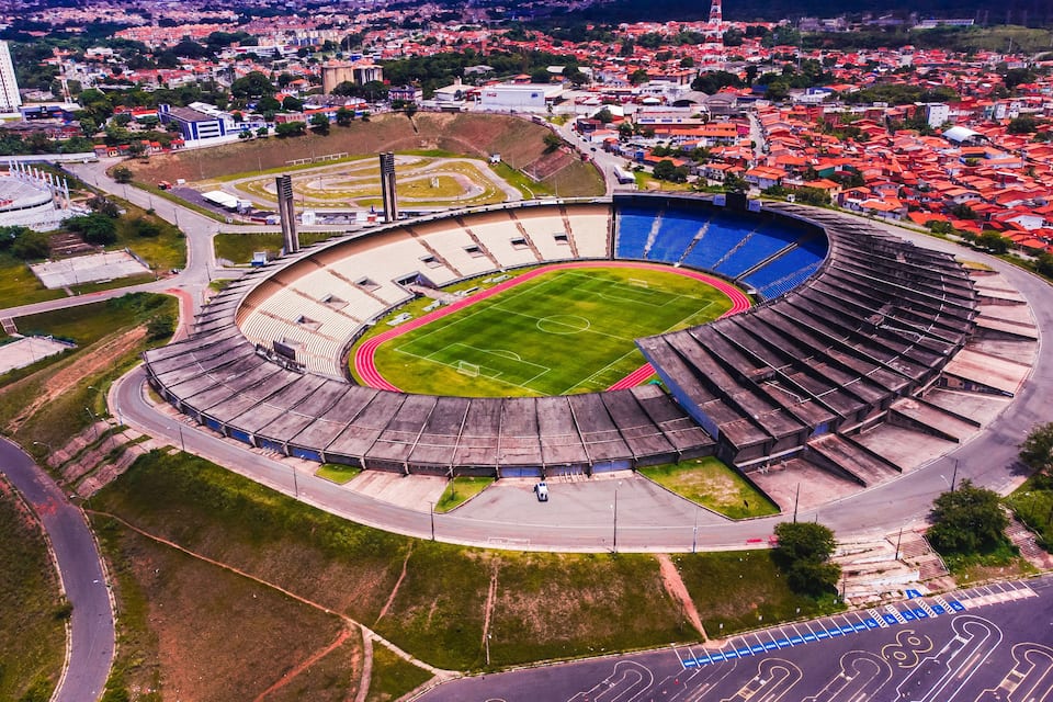 Vista aérea de um estádio grande com campo de futebol, cercado por uma paisagem urbana com casas de telhado vermelho.