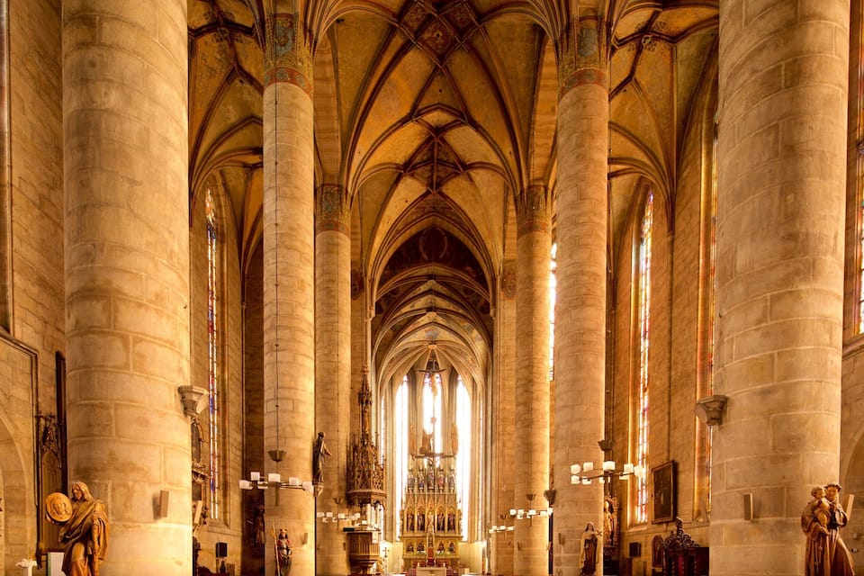 Interior de uma catedral com tetos abobadados altos, grandes vitrais e estátuas.