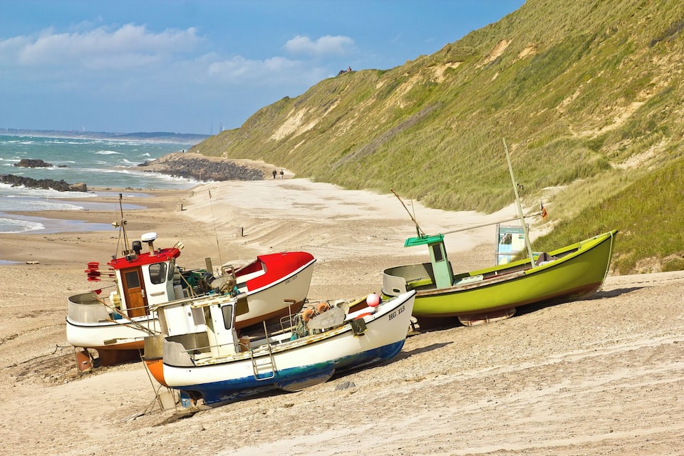 Uma praia com três barcos, areal arenoso e uma colina verdejante ao fundo.
