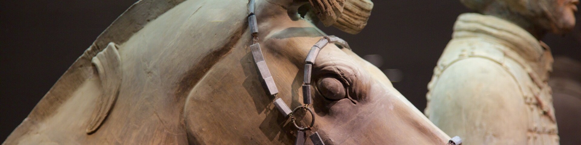 Close-up de uma estátua de cavalo com freio detalhado e uma figura em armadura ao fundo.