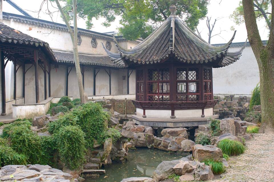 Um pavilhão chinês tradicional em um jardim com um pequeno lago e terreno rochoso.