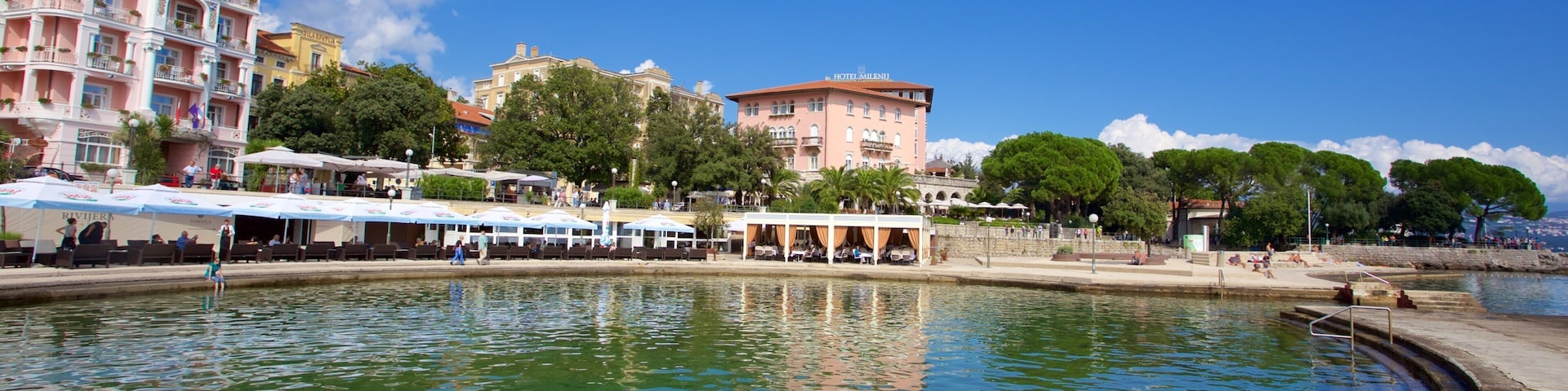 Um calçadão à beira-mar com céu azul claro, um hotel e um restaurante com mesas ao ar livre.