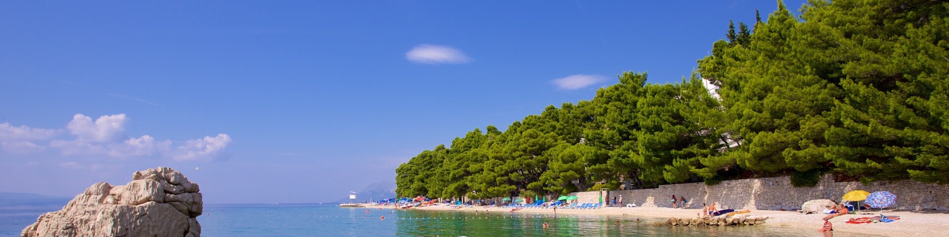 Mar turquesa e translúcida, costa rochosa e uma praia com guarda-sóis e pessoas.
