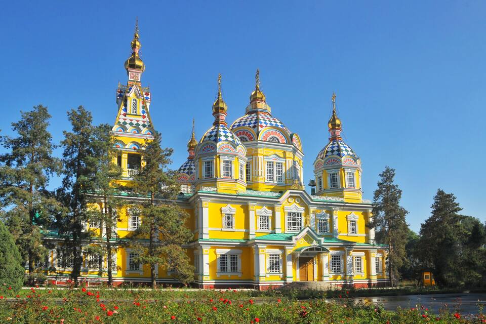 Uma igreja colorida com várias cúpulas e telhado verde, cercada por árvores e flores vermelhas.