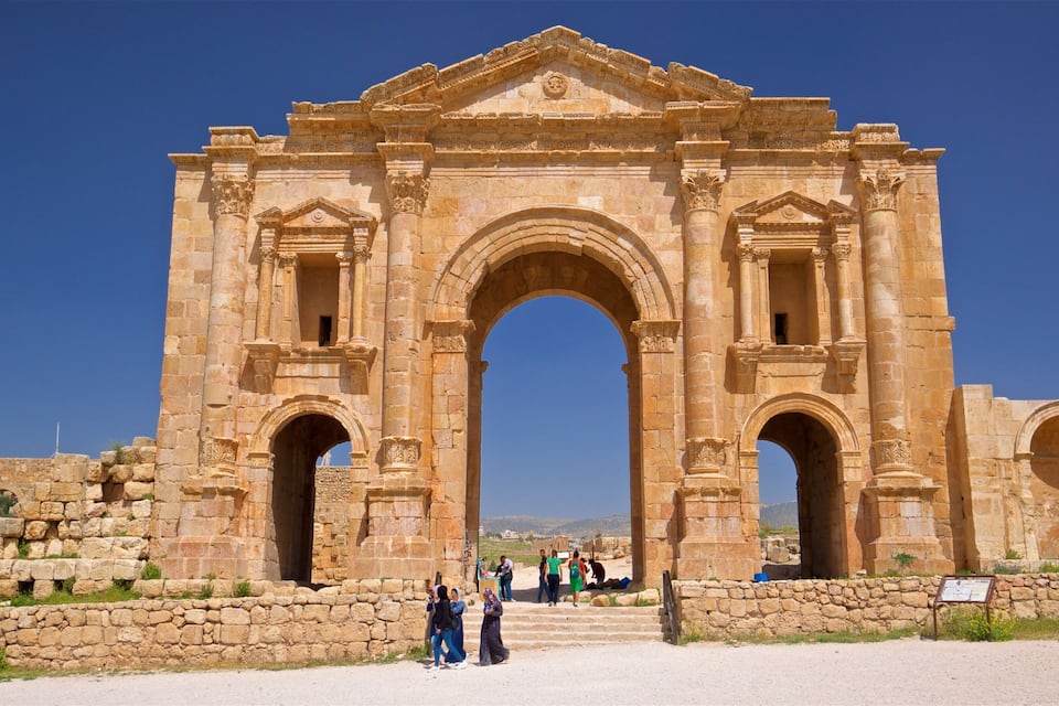 Um arco de pedra antiga com esculturas detalhadas e colunas, cercado por visitantes e um céu azul límpido.