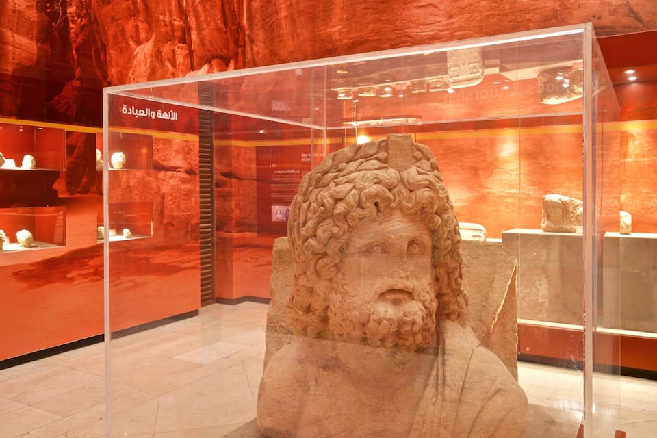 Uma escultura de pedra em um museu, com paredes vermelhas e vitrines de vidro.