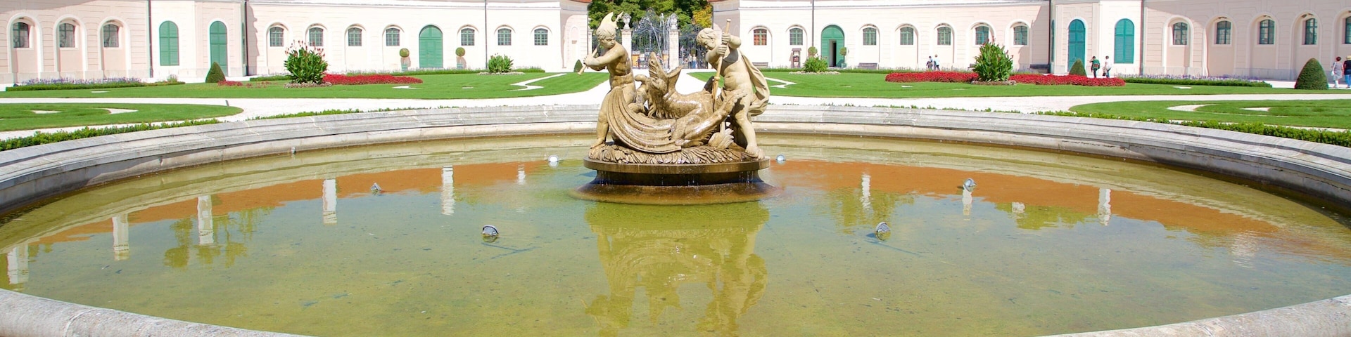 Uma fonte clássica com estátuas num jardim formal, rodeada por relvados bem cuidados e flores vermelhas.