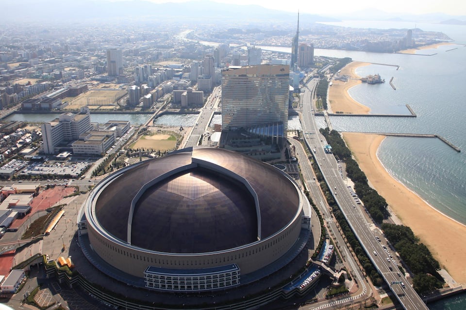 Paisagem costeira com um estádio grande, edifícios altos e uma praia.