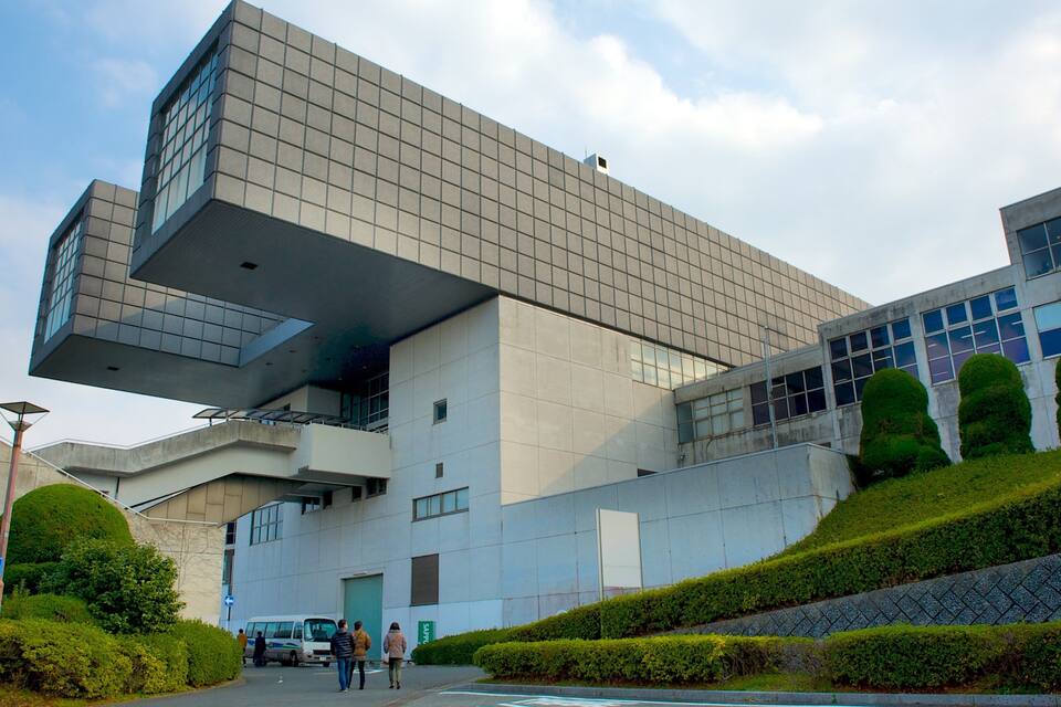 Um edifício de arquitetura moderna com uma seção em balanço singular, cercado por sebes bem cuidadas e um céu limpo.