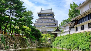 Um castelo japonês tradicional com um lago em primeiro plano, rodeado por vegetação exuberante e muros de pedra.