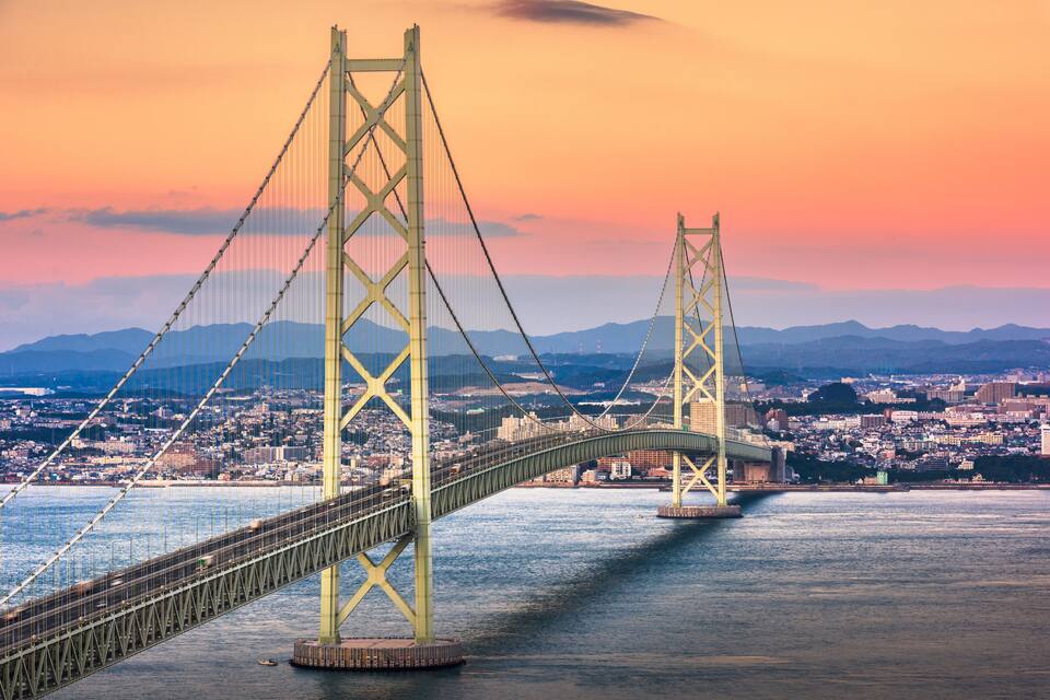 Uma ponte suspensa com duas torres, atravessando um corpo de água, com um horizonte urbano ao fundo.