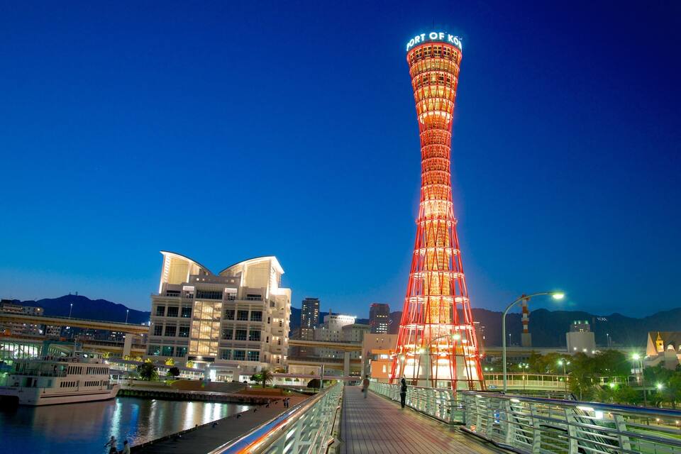 Uma torre bem iluminada com o texto “Port of Kochi” no topo, um edifício moderno com fachada curva, e um rio com uma ponte em primeiro plano.