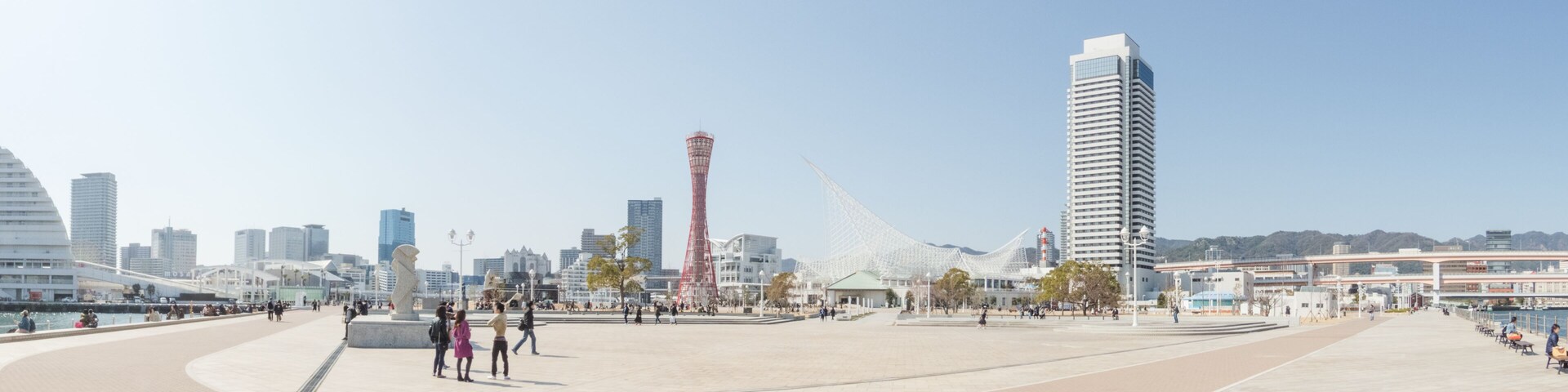 Uma ampla praça pública com arquitetura moderna, incluindo uma torre vermelha alta e um edifício branco com telhado curvo.
