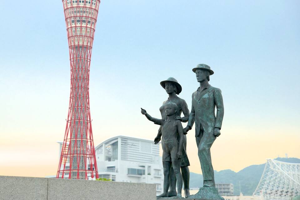 Estátua de um homem, uma mulher e uma criança, uma torre vermelha e edifícios modernos ao fundo.