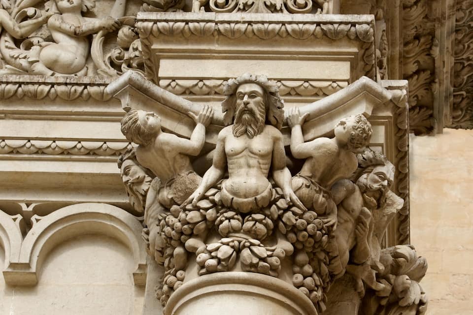Detalhada escultura em pedra de uma figura sentada, rodeada por figuras menores e elementos decorativos.