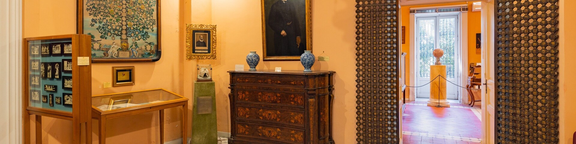 Um corredor com ladrilhos estampados, um quadro grande emoldurado, um armário de madeira e uma porta com grade decorativa.