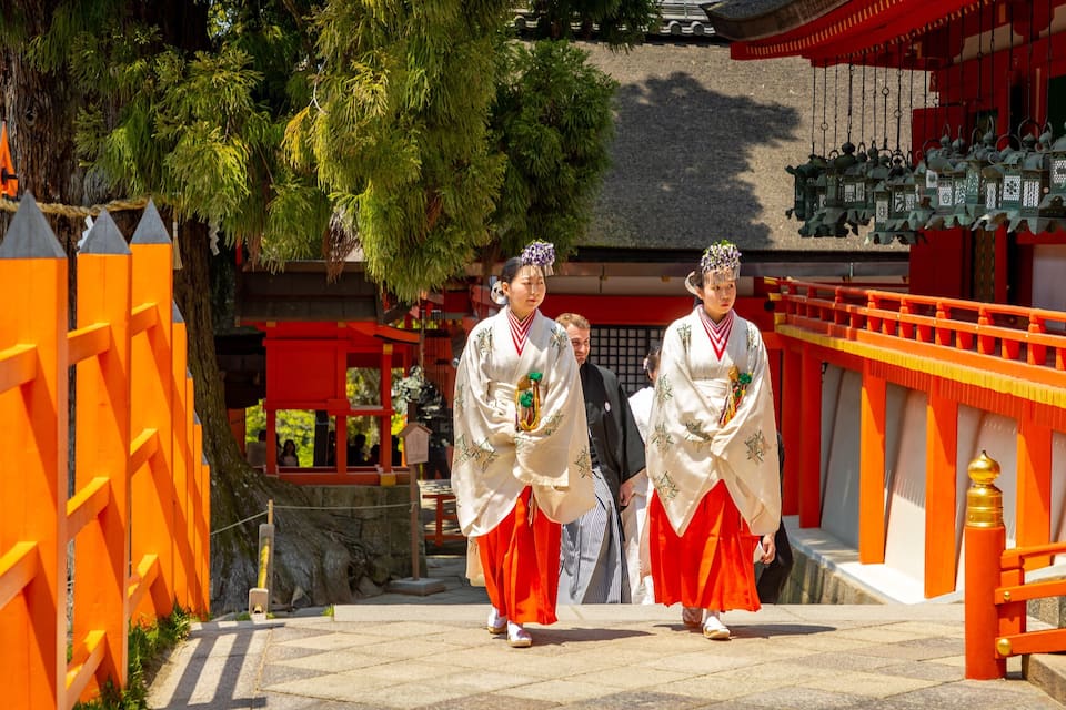 Um santuário japonês tradicional com estruturas em laranja e vermelho, um caminho de pedra e duas pessoas vestidas com trajes tradicionais.
