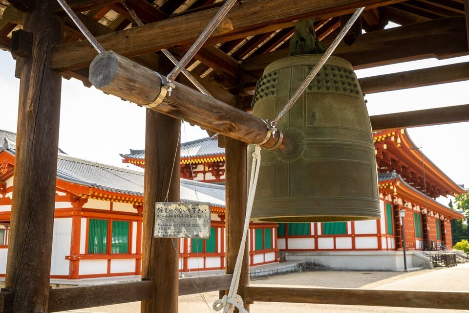 Um sino grande suspenso em uma estrutura de madeira, com arquitetura japonesa tradicional ao fundo.