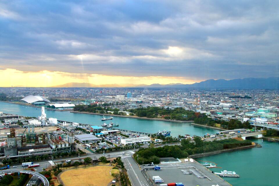 Paisagem urbana com um rio, área industrial, estádio e montanhas ao fundo.