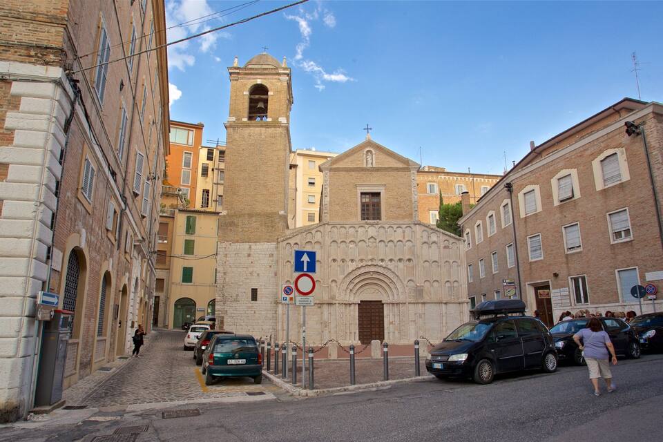 Uma rua de paralelepípedos com edifícios históricos, uma igreja com campanário e carros estacionados.
