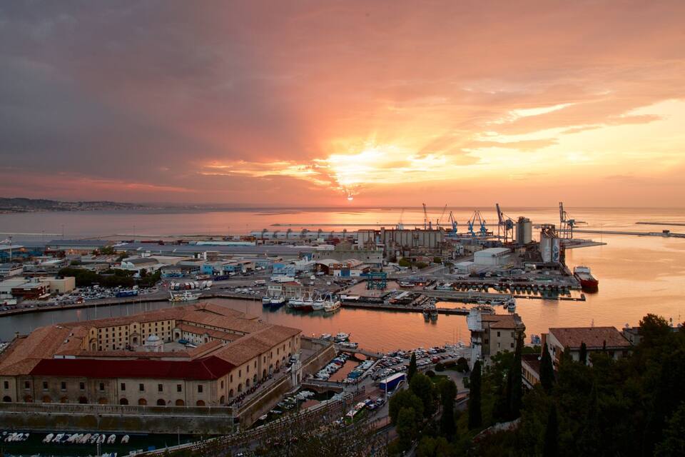 Paisagem costeira ao pôr do sol, com um porto importante, edifícios históricos e vista clara do mar.