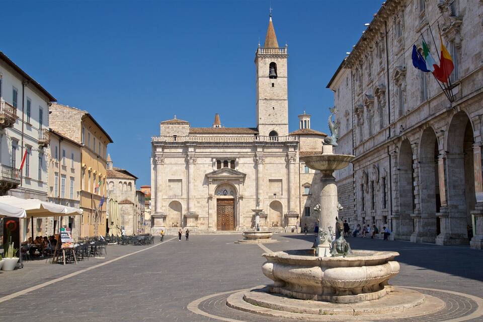 Uma praça histórica com uma igreja imponente, um campanário, uma fonte e edifícios ao redor com bandeiras.