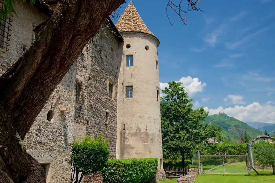 Uma torre histórica com telhado cónico, rodeada por vegetação exuberante e montanhas ao fundo.