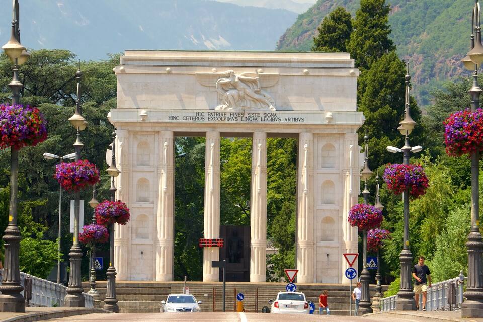 Um monumento neoclássico imponente com inscrições, rodeado por vegetação exuberante e montanhas ao fundo.