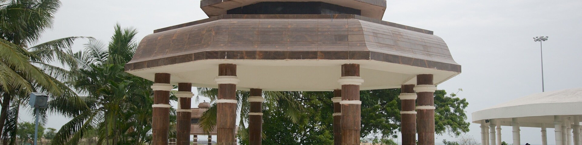 Um pavilhão com telhado abobadado e base de pedra, rodeado por palmeiras e um muro branco.
