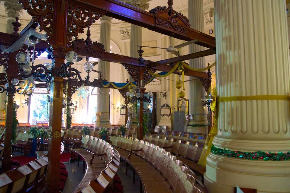 Interior de igreja ricamente decorado, com fileiras de bancos, uma coluna branca imponente e lustres ornamentais.