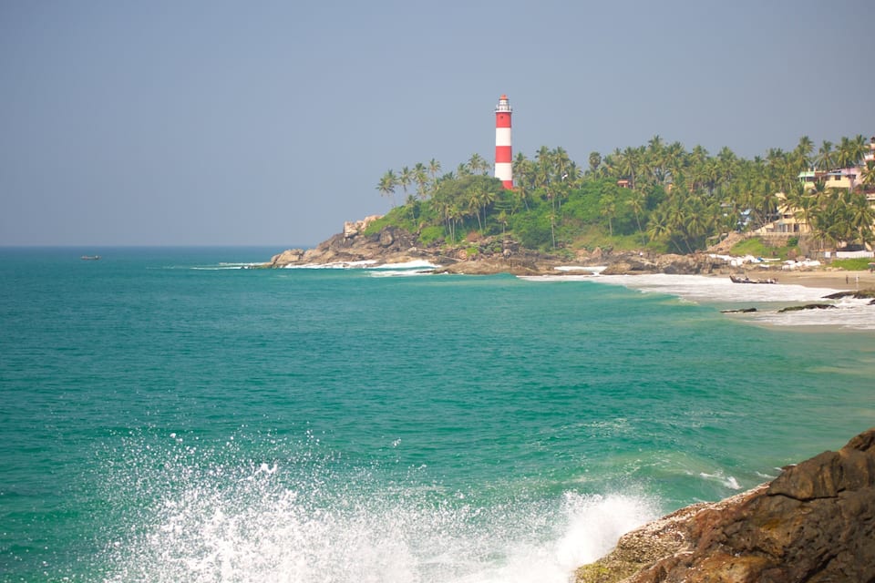 Cena costeira com um farol numa falésia rochosa, palmeiras e uma praia com edifícios ao fundo.